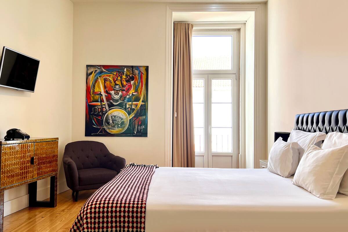 Quarto de hotel com cama, cadeira, escrivaninha, televisão e um quadro na parede.