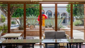 Uma área de refeições interna com grandes janelas, vista para o jardim externo e uma escultura vermelha.