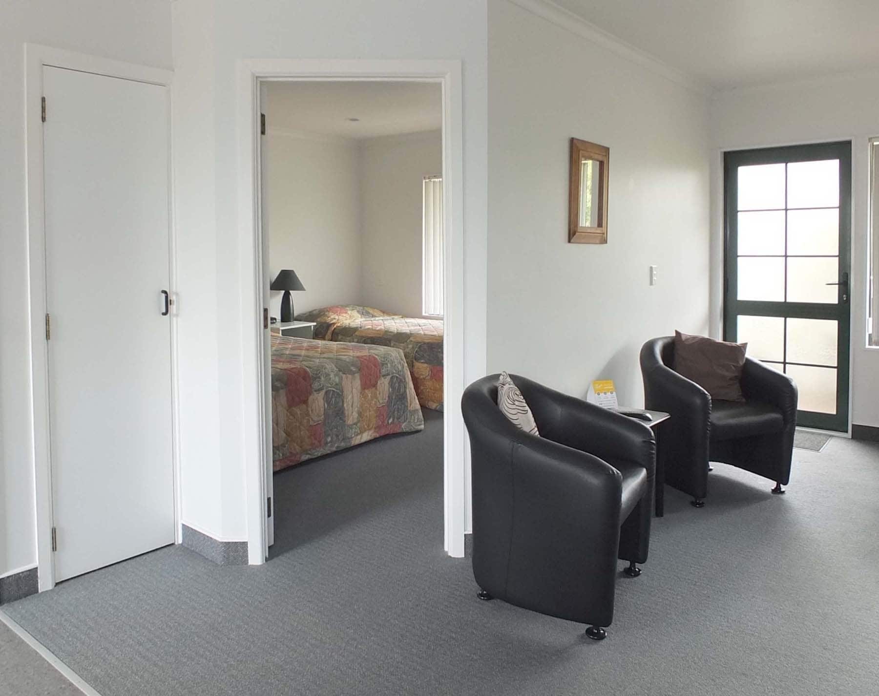 Quarto de hotel com uma cama, duas cadeiras, uma mesa de trabalho e uma porta que dá para outro cômodo.