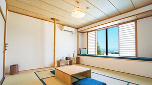 Um quarto japonês tradicional com tatames, uma mesa de madeira baixa, um travesseiro azul e uma grande janela com vista para o mar.