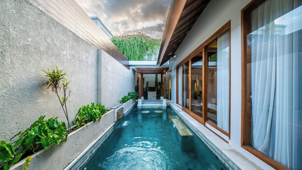 Uma piscina estreita com borda de pedra, ladeada por um muro de concreto e uma pérgola de madeira.