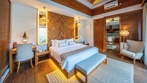 Quarto de hotel moderno com teto de madeira, uma cama grande com colcha azul, uma escrivaninha com luminária e um espaço de estar com poltrona e banco.