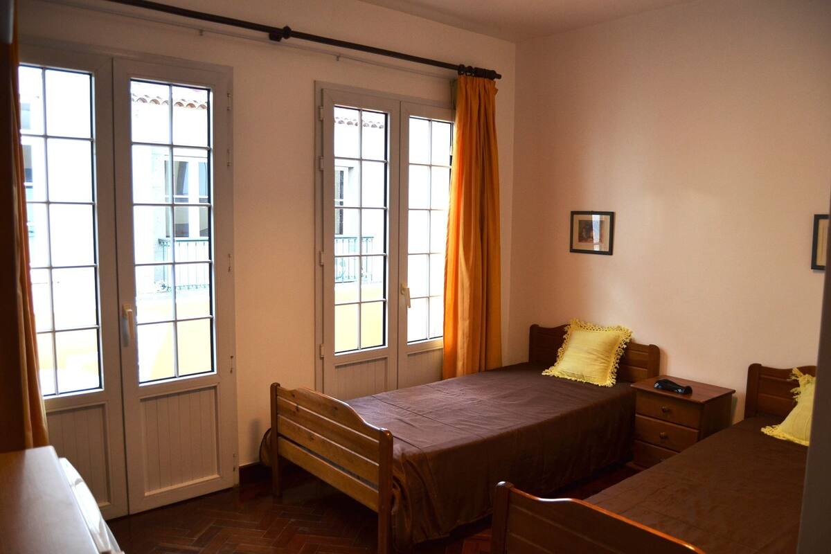 Quarto com duas camas, um banco de madeira, uma mesinha de cabeceira e uma janela com cortinas.