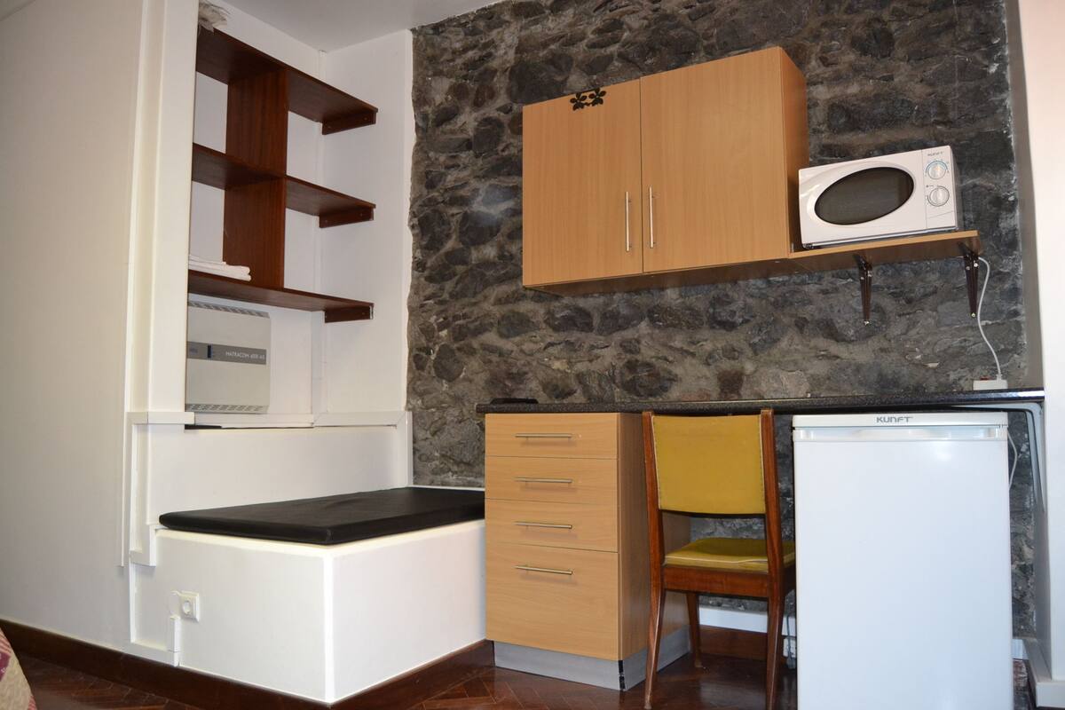Uma cozinha compacta com parede de pedra, armários de madeira, micro-ondas, mini-geladeira e uma cadeira amarela.