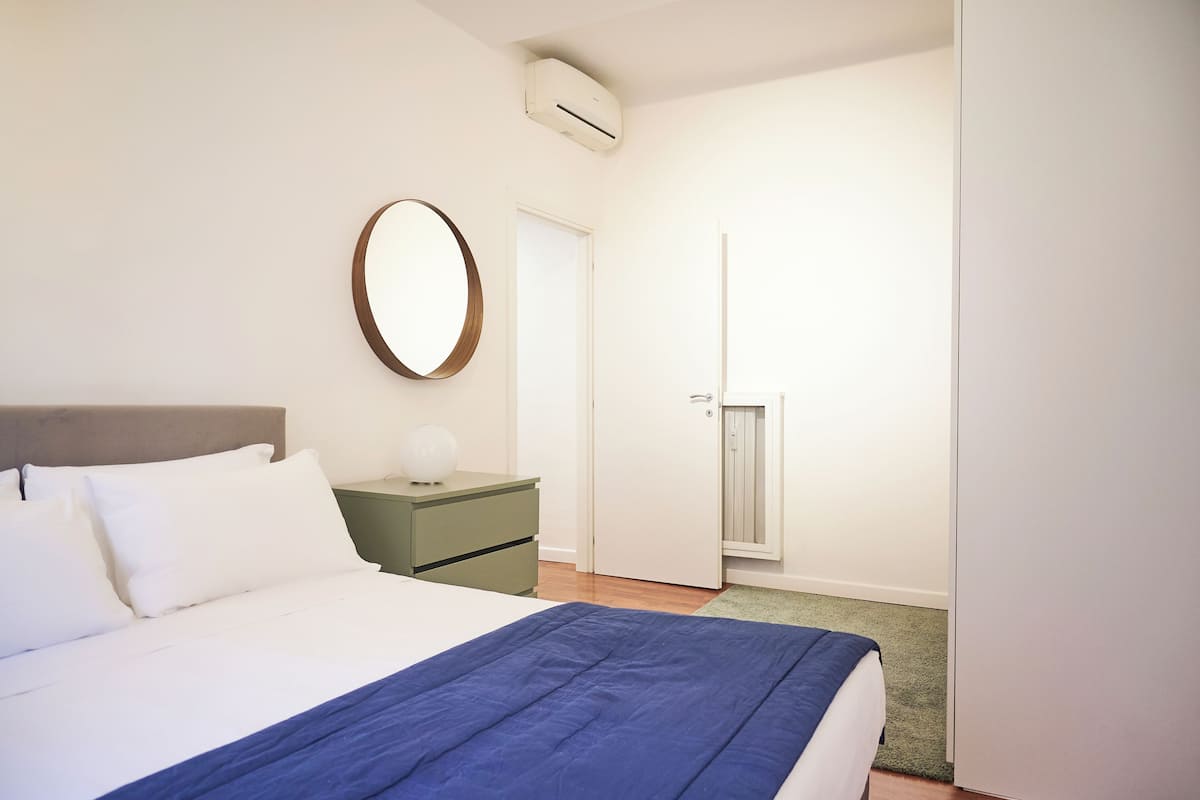 Um quarto com uma cama, mesa de cabeceira, espelho e porta.