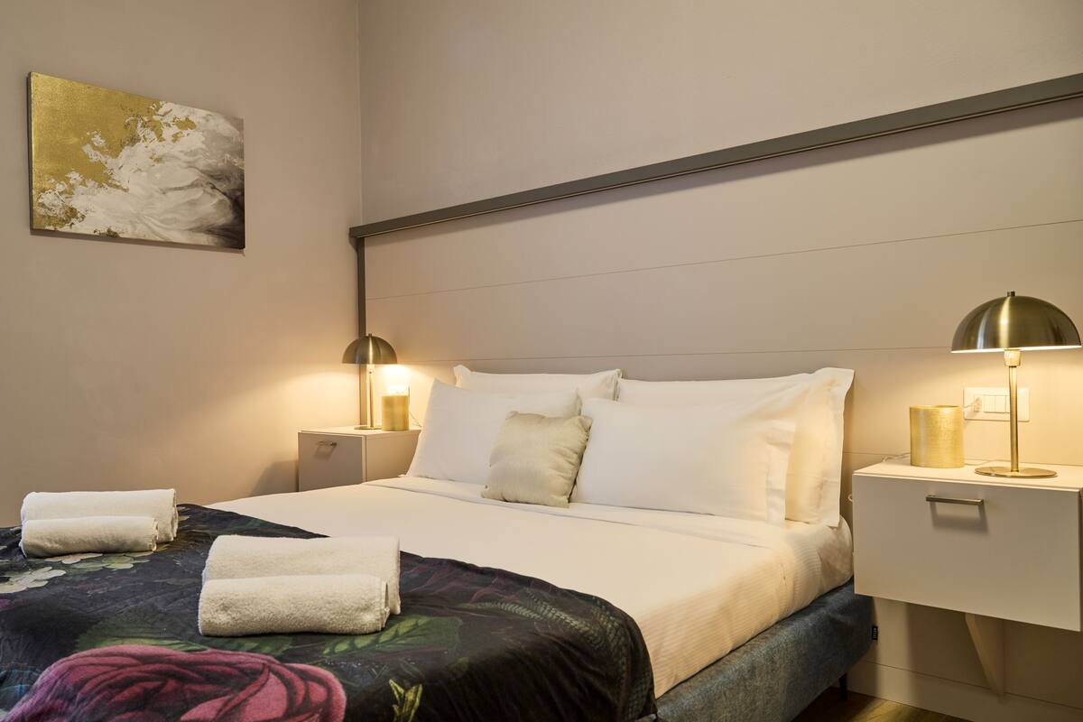 Cama bem arrumada com lençóis brancos, um cobertor estampado com motivos florais e duas toalhas enroladas ao lado. Há duas luminárias de cabeceira e uma pequena mesa de cabeceira com um abajur.