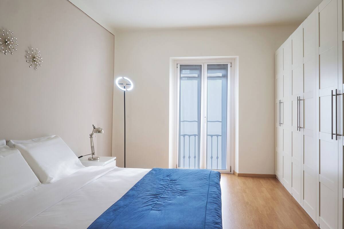 Quarto com cama, luminária de cabeceira, guarda-roupa e janela com varanda.