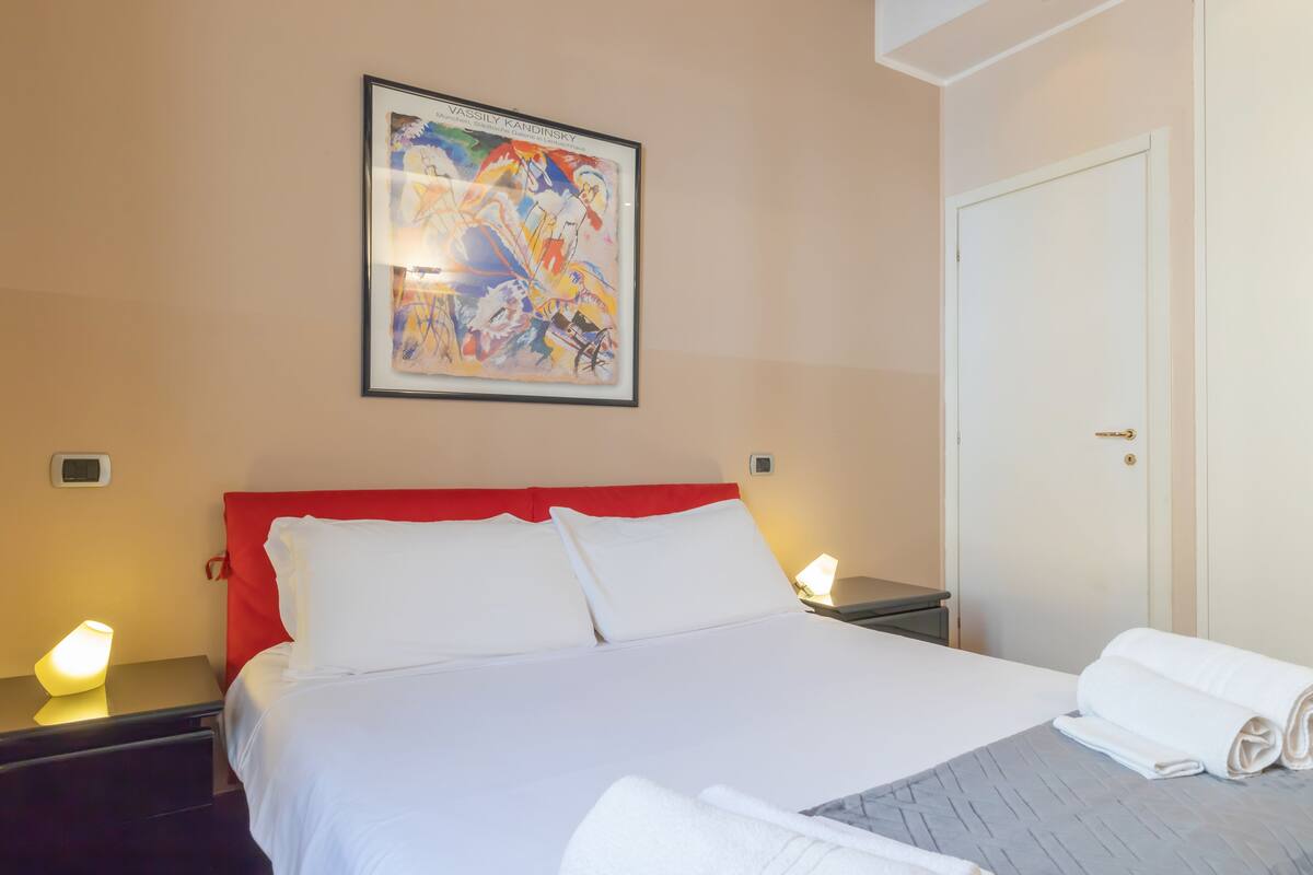Quarto de hotel com cama, mesa de cabeceira, quadro na parede, porta e relógio de parede.