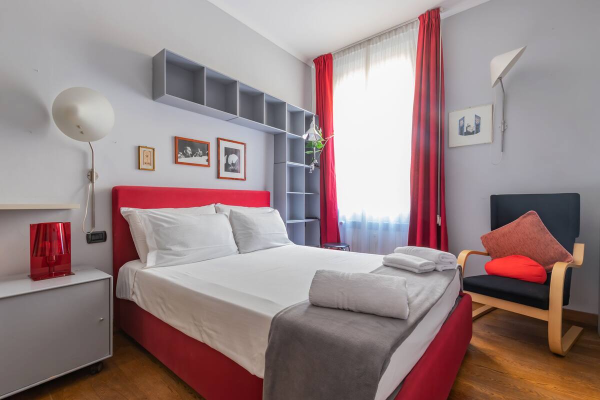 Quarto de hotel moderno com cama vermelha, prateleiras cinzas, cadeira vermelha e mesa de cabeceira branca.