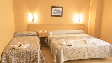 Quarto de hotel com duas camas, cada uma com um edredom estampado e uma toalha dobrada. Há uma mesa de cabeceira entre as camas, com uma luminária e uma pequena mesa.