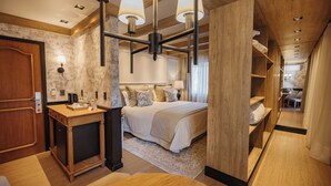 Quarto de hotel moderno com piso de madeira, uma cama grande com colcha bege, mesa de cabeceira de madeira com abajur e guarda-roupa de madeira com prateleiras.