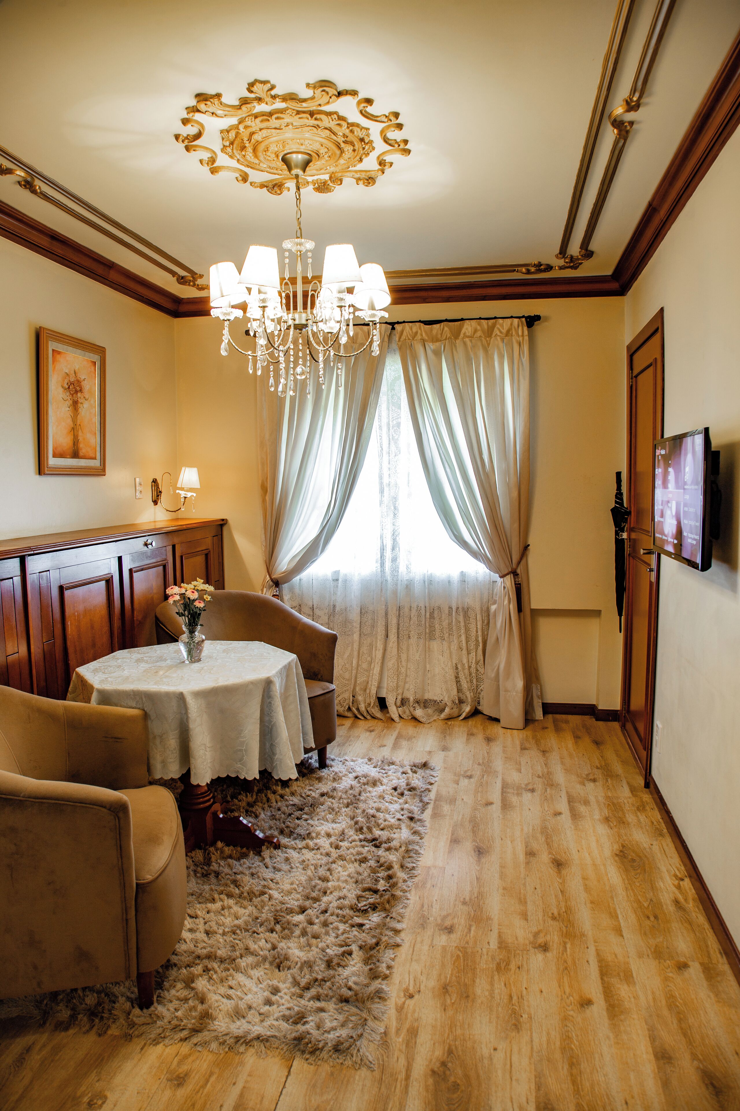 Quarto com piso de madeira, um candelabro, uma mesa redonda com um vaso de flores, duas poltronas, uma televisão na parede e uma janela com cortinas.