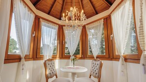 Quarto com teto de madeira, um candelabro, duas poltronas estofadas, uma mesa redonda com um vaso e grandes janelas com cortinas.