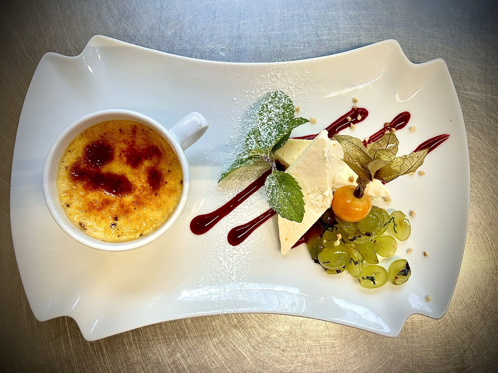 Um prato de sobremesa com crème brûlée, uvas, queijo e elementos decorativos.