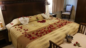 Cama bem arrumada com pétalas de rosa e velas, mesa de cabeceira de madeira, uma cadeira e uma mesa com um balde e uma garrafa.