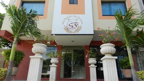 Entrada de hotel com placa central, duas colunas imponentes e plantas em vasos.