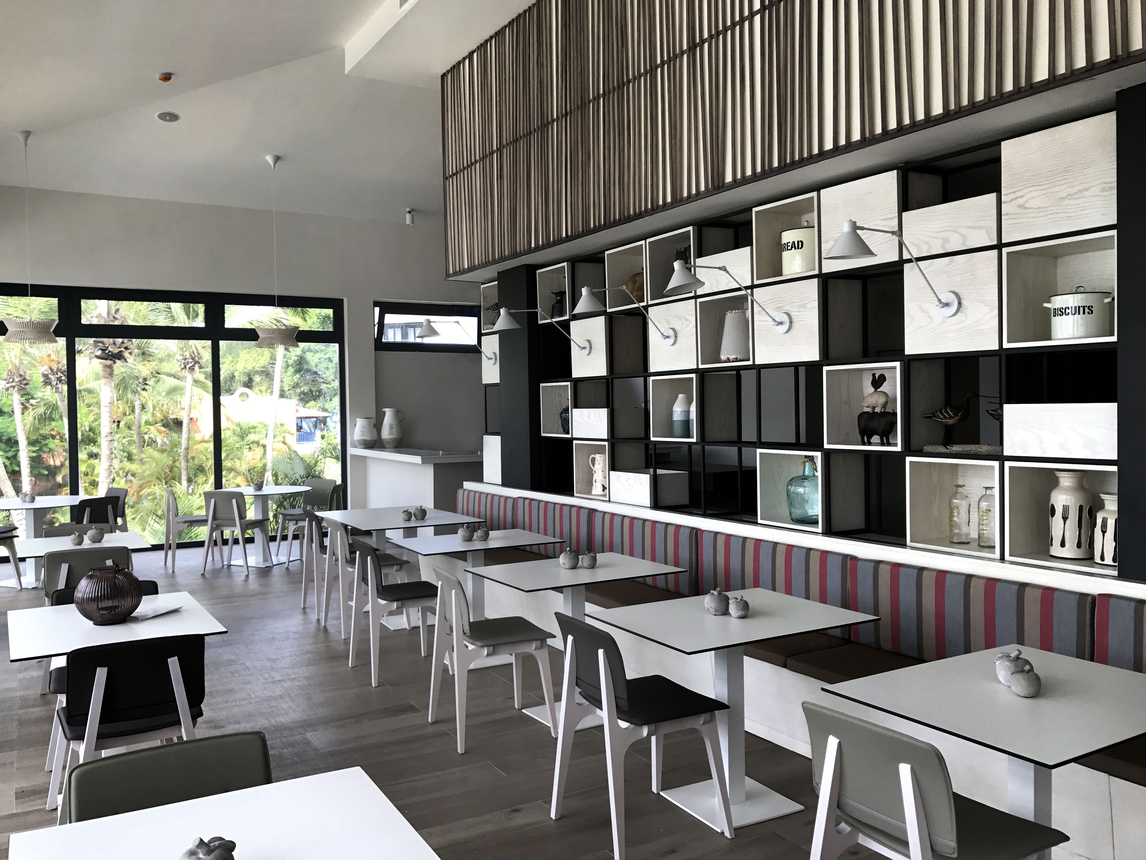 Um café moderno com mesas e cadeiras brancas, um banco riscado e uma parede com caixas e prateleiras decorativas.