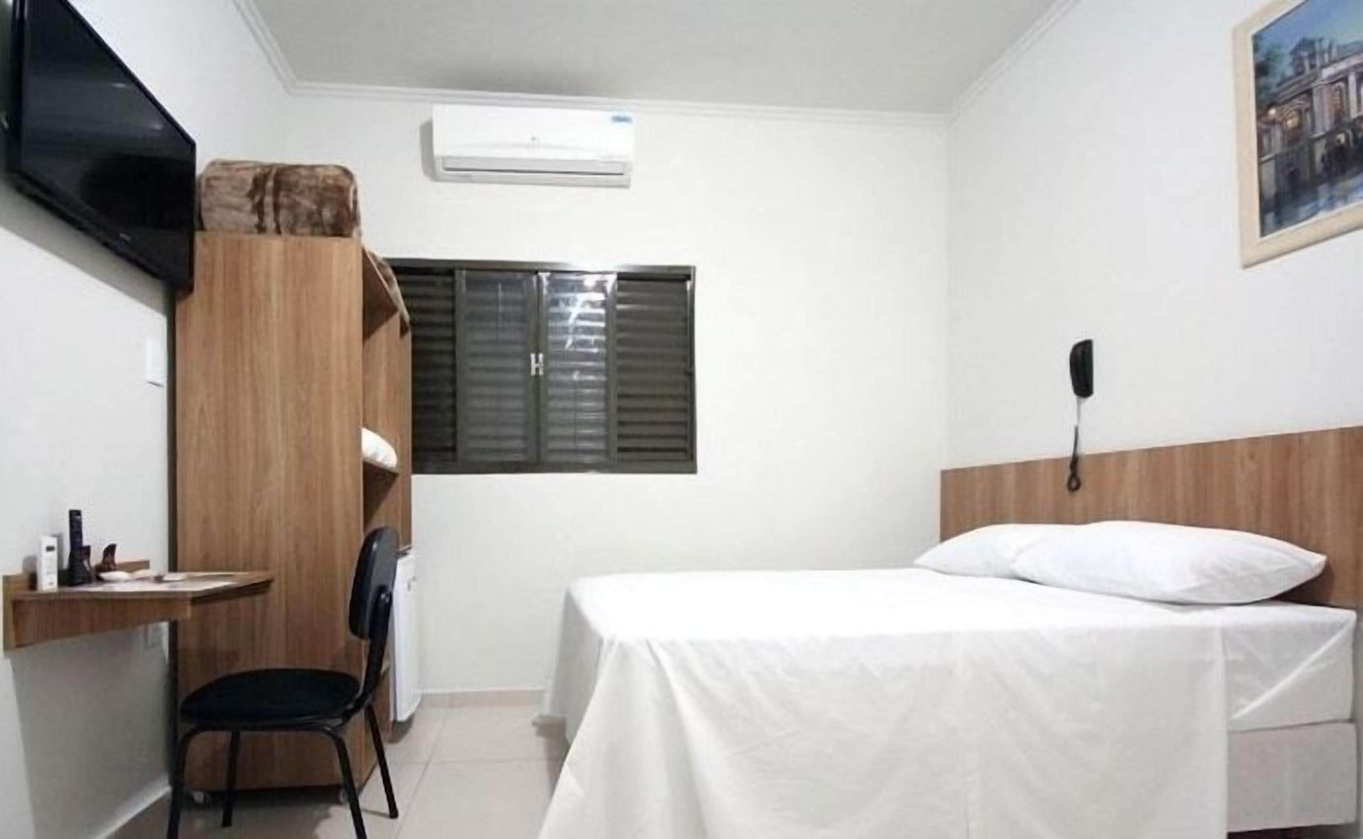 Quarto pequeno e limpo com cama, escrivaninha, cadeira, televisão e janela com persianas.