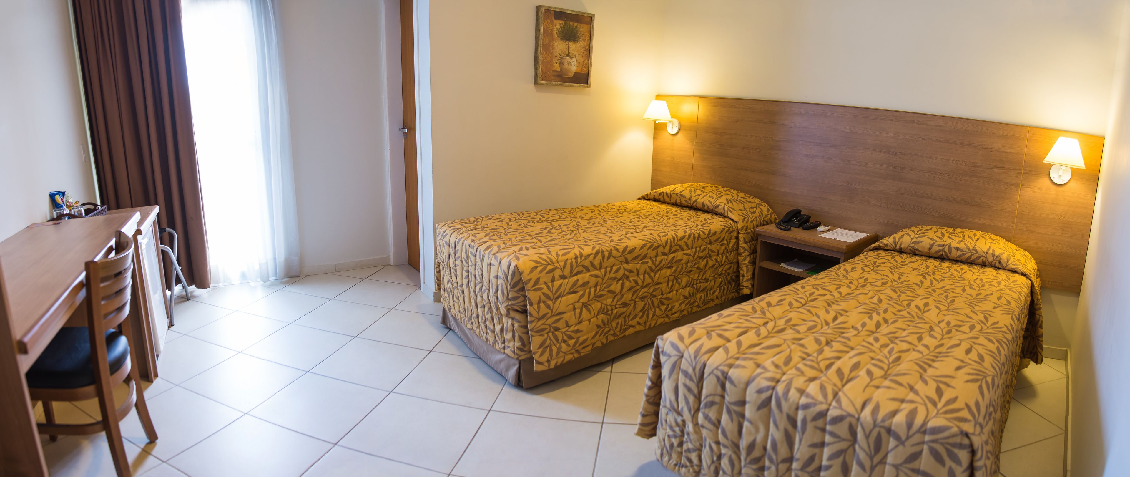 Quarto de hotel com duas camas de solteiro, uma escrivaninha com cadeiras, um telefone e uma luminária na parede.