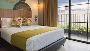 Quarto de hotel moderno com uma cama grande, cabeceira de madeira, manta verde e vista para a cidade por amplas janelas.