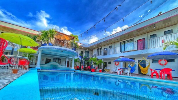 Uma área de piscina com uma piscina azul cristalina, cadeiras de sol vermelhas e azuis, e guarda-sóis.