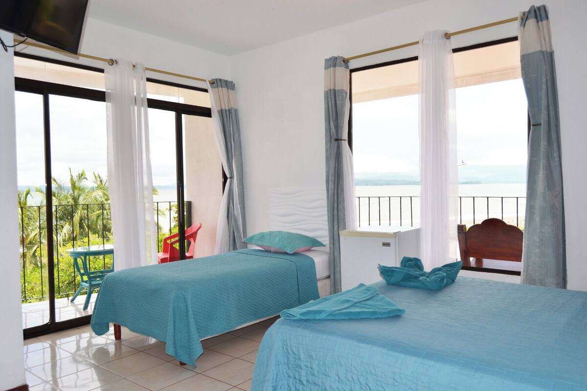 Quarto de hotel com duas camas, varanda com mesa e cadeiras, e vista para o mar.