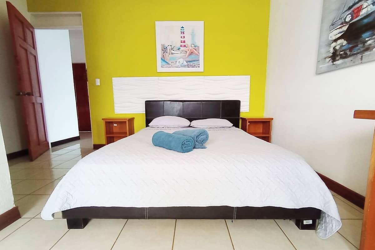 Quarto de hotel com uma cama grande, roupa de cama branca e parede amarela.