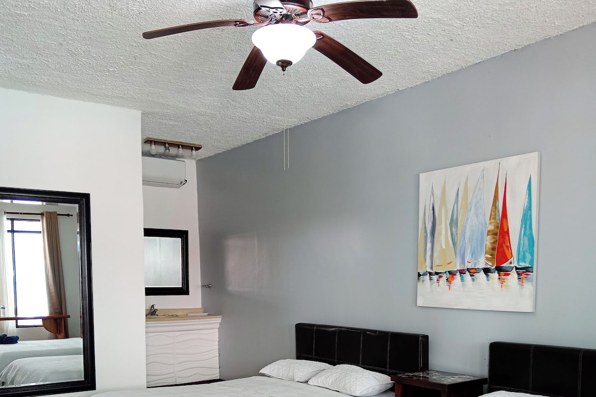 Quarto de hotel com duas camas, um ventilador de teto, pintura de barcos à vela, um espelho grande e uma janela com cortinas.
