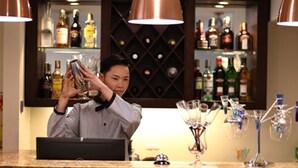 Uma área de bar com um bartender preparando uma bebida, um laptop no balcão e várias garrafas ao fundo.