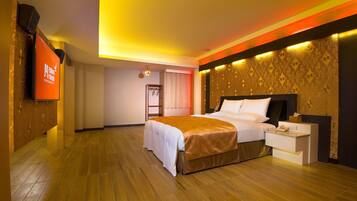 Quarto de hotel com uma cama grande, uma mesinha de cabeceira, televisão fixada na parede e piso de madeira.