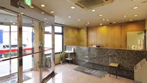 Um hall de hotel com entrada por porta de vidro, balcão de recepção, planta em vaso e um pequeno banquinho.