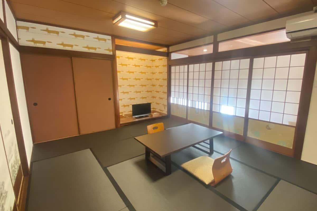 Um quarto japonês tradicional com tatame, uma mesa baixa, uma televisão, portas de correr e vista através de um biombô shoji.