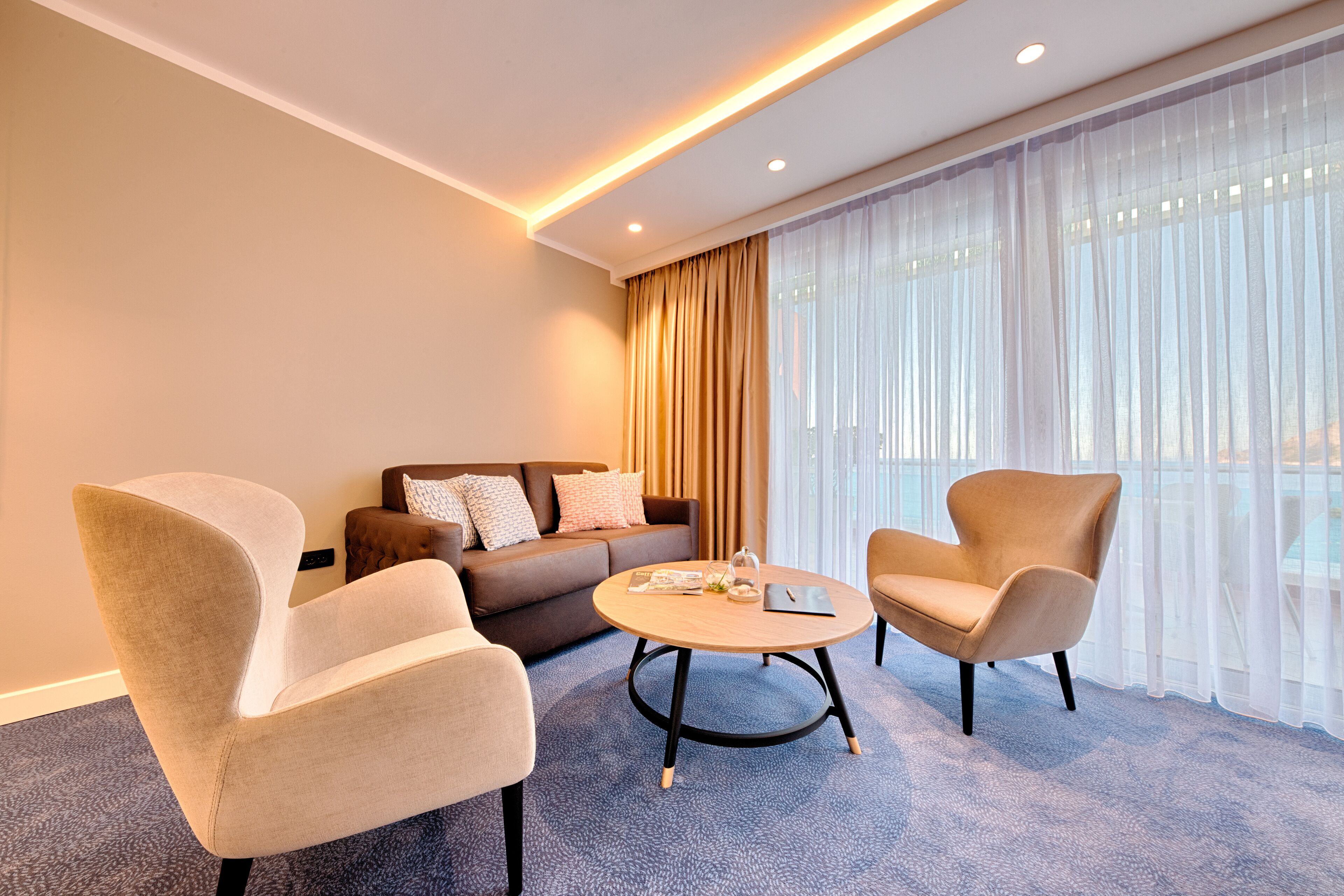 Quarto de hotel moderno com sofá, duas poltronas, mesa de centro redonda e uma janela grande com cortinas leves.