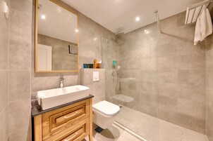 Uma casa de banho moderna com um espelho grande, uma sanita, um lavatório branco, um toucador de madeira e uma cabine de duche com parede de vidro.