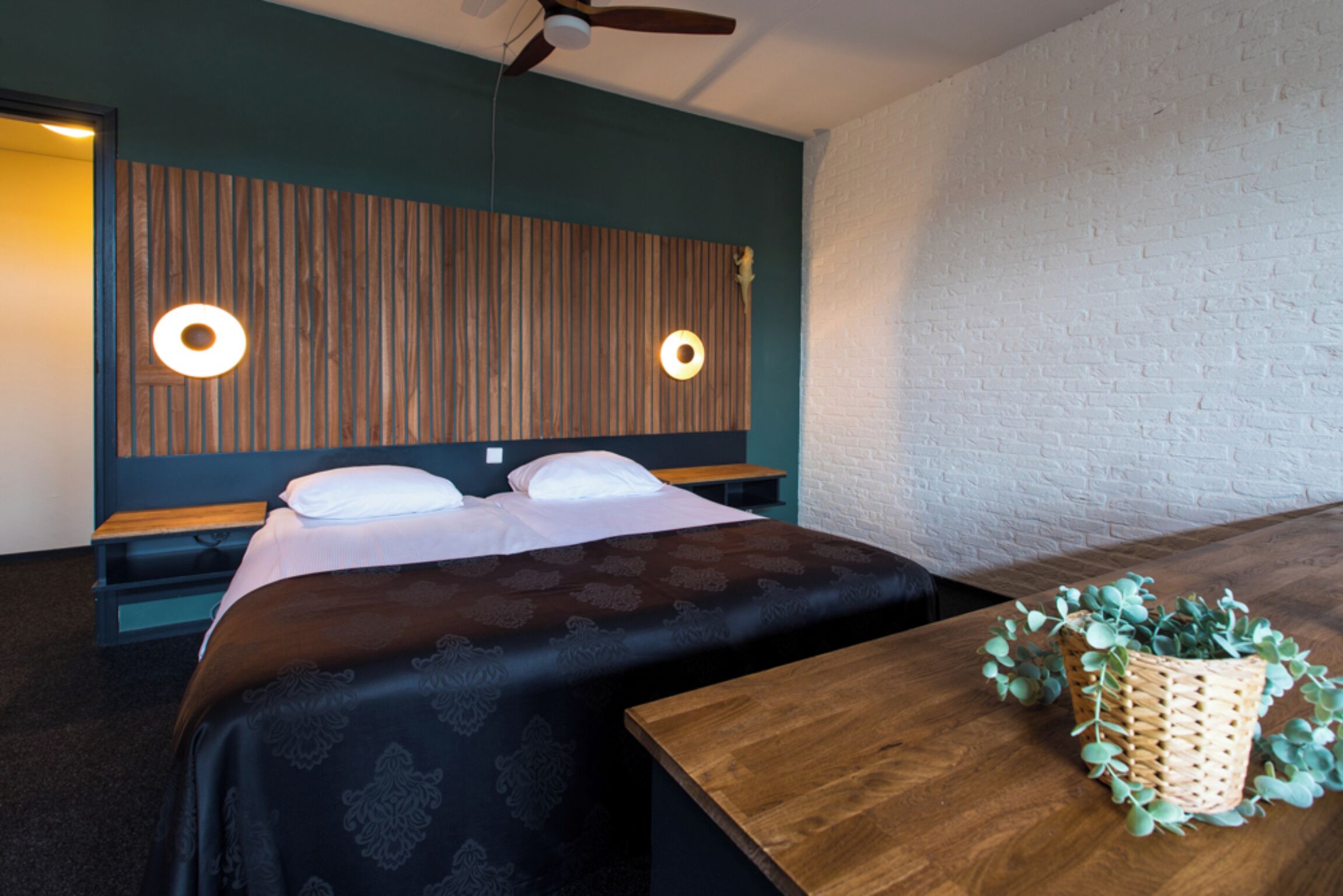 Quarto de hotel moderno com cabeceira de madeira, cama com colcha estampada escura, mesa de cabeceira de madeira, mesa e cesta de vime com folhagem.