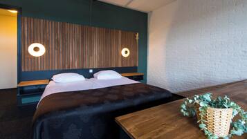 Quarto de hotel moderno com cabeceira de madeira, cama com colcha estampada escura, mesa de cabeceira de madeira, mesa e cesta de vime com folhagem.