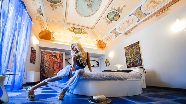 Quarto com cama redonda, uma pintura grande e teto com detalhes ornamentados.