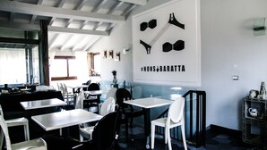 Interior de restaurante com mesas e cadeiras, uma parede com o letreiro #NONSOBARATTA e vista para o exterior através de uma janela.