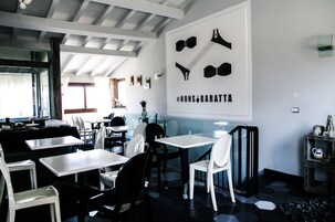 Interior de restaurante com mesas e cadeiras, uma parede com o letreiro #NONSOBARATTA e vista para o exterior através de uma janela.