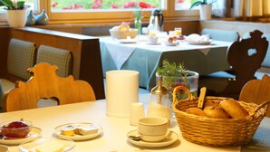 Uma área de refeições com uma mesa posta para o café da manhã, incluindo pão, manteiga e geleia. Há cadeiras e um banco ao redor da mesa, e uma janela com vista para o verde lá fora.