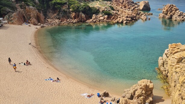 Uma praia com águas turquesa cristalinas, falésias rochosas e uma escadaria de madeira que desce até a costa.