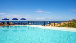 Piscina com águas cristalinas azuis, rodeada por espreguiçadeiras e guarda-sóis azuis, com vista para a costa rochosa.