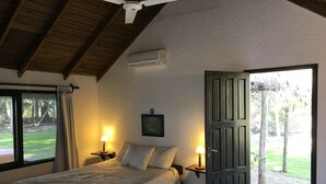 Quarto com cama, duas luminárias de cabeceira, ventilador de teto e porta aberta para área externa.