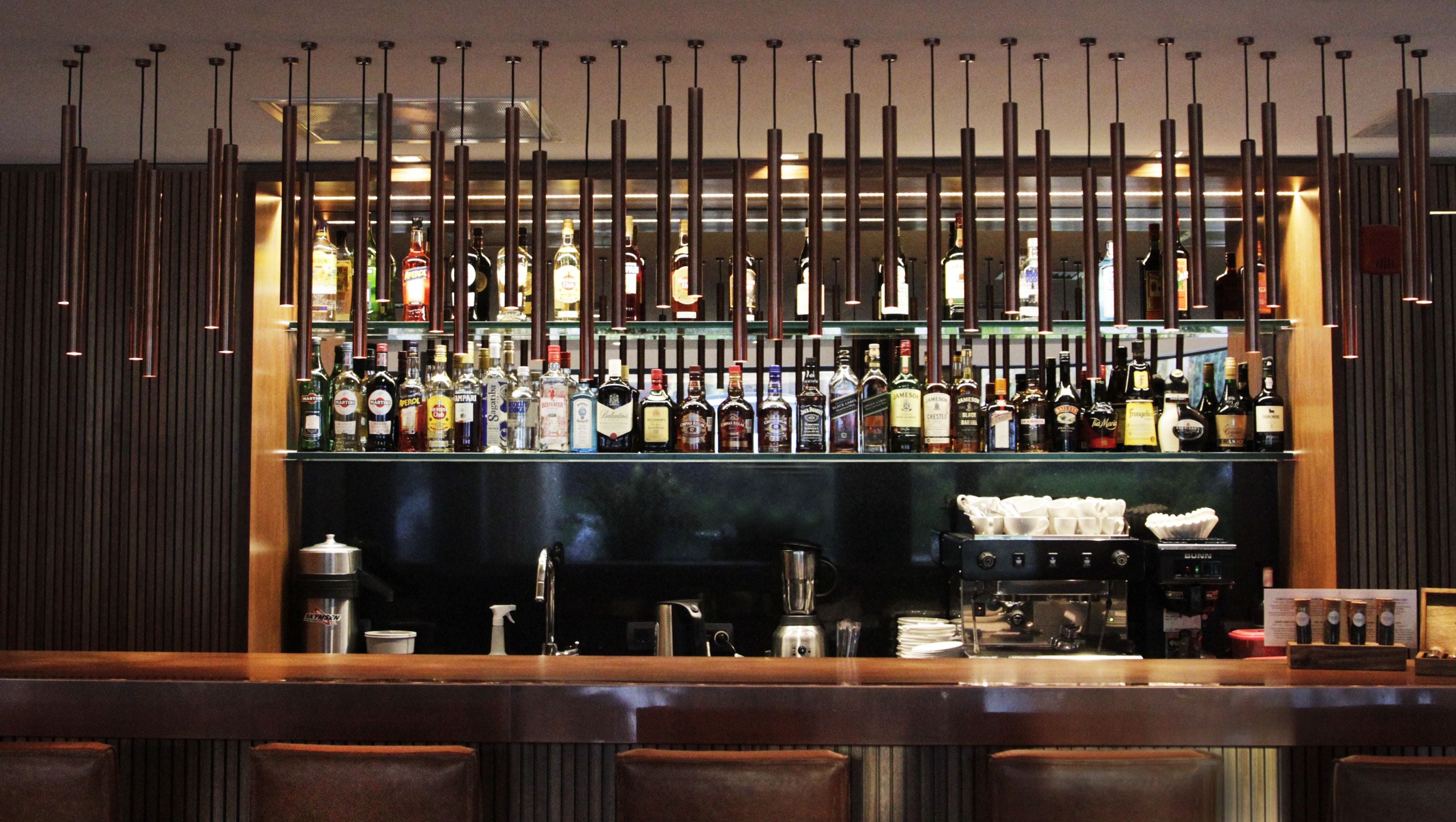 Uma área de bar com diversas garrafas de bebidas, uma máquina de café e um conjunto de copos brancos.