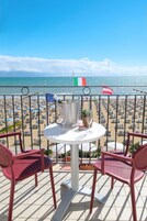 Varanda com mesa e cadeiras, vista para uma praia com guarda-sóis e bandeiras.