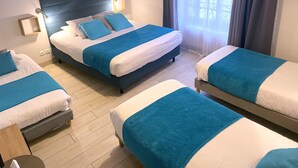 Quarto de hotel com três camas, cada uma com roupa de cama azul e branca, piso de madeira e uma grande janela com cortinas.