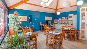 Uma área de refeições com mesas e cadeiras de madeira, uma máquina de café e vista para o exterior através de uma janela.