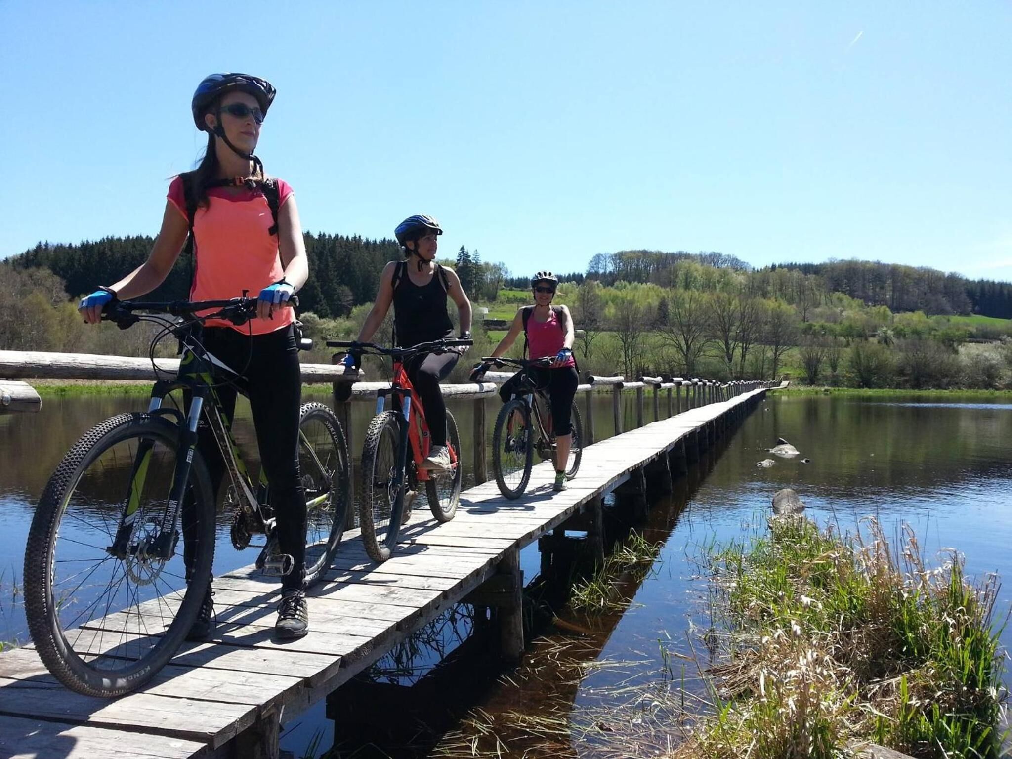 Três ciclistas em uma ponte de madeira sobre um lago calmo, cercados por vegetação e árvores.