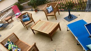 Uma varanda com mobiliário de madeira, incluindo cadeiras, uma mesa e uma poltrona, com vista para um jardim e uma grade de metal.