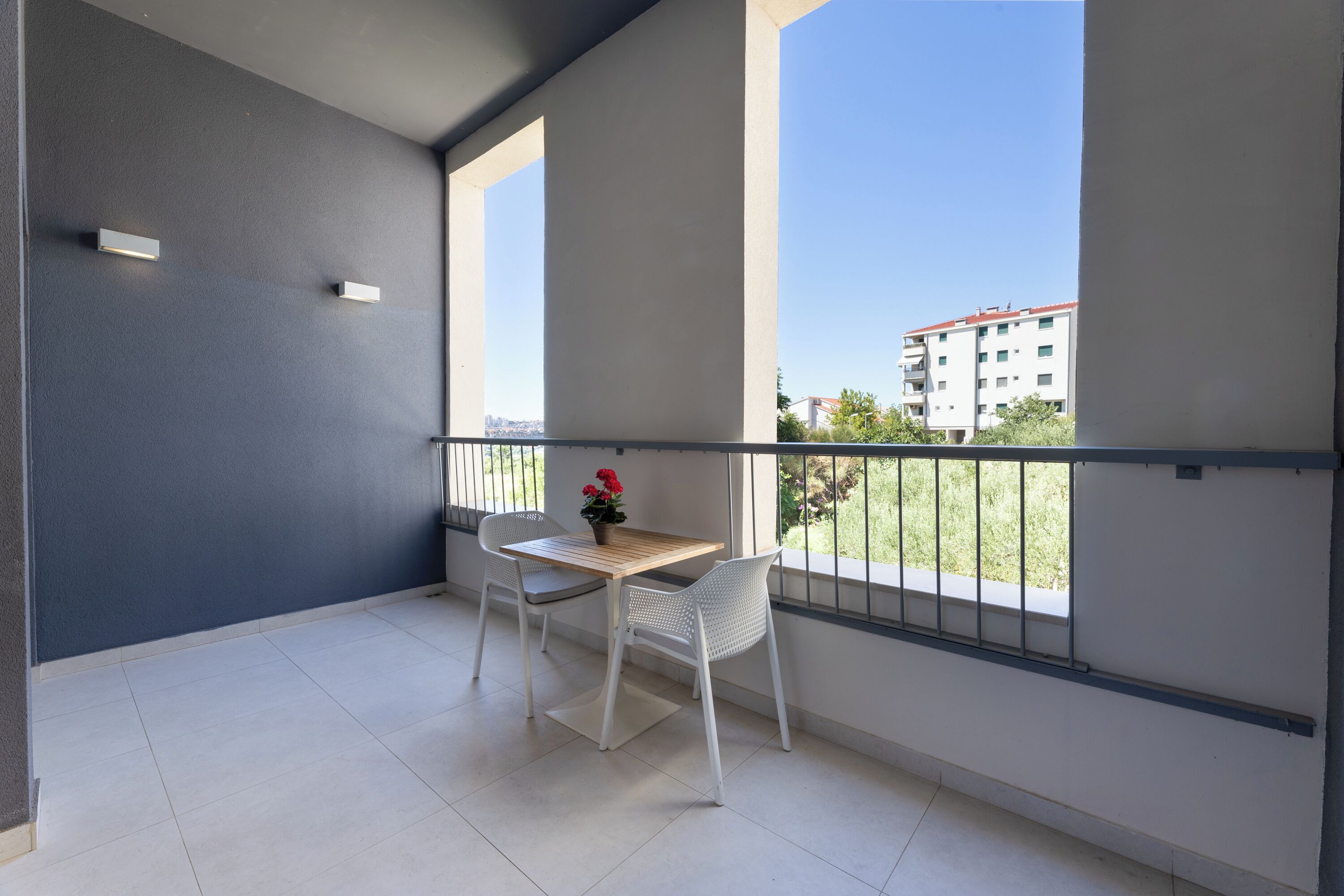 Uma varanda com mesa e duas cadeiras, um vaso de planta e vista para um edifício ao fundo.